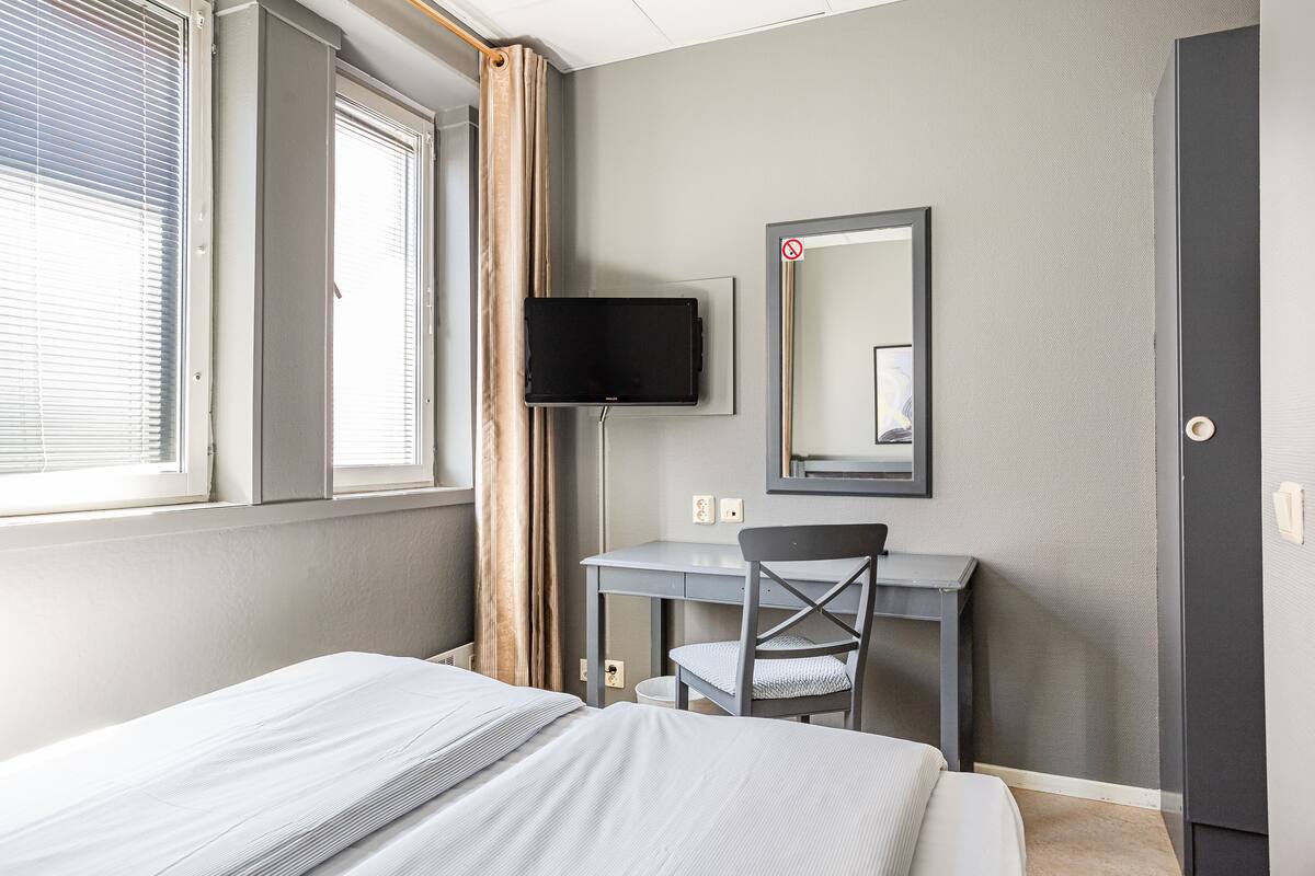 Quarto de hotel com cama, escrivaninha, cadeira, televisão e espelho.