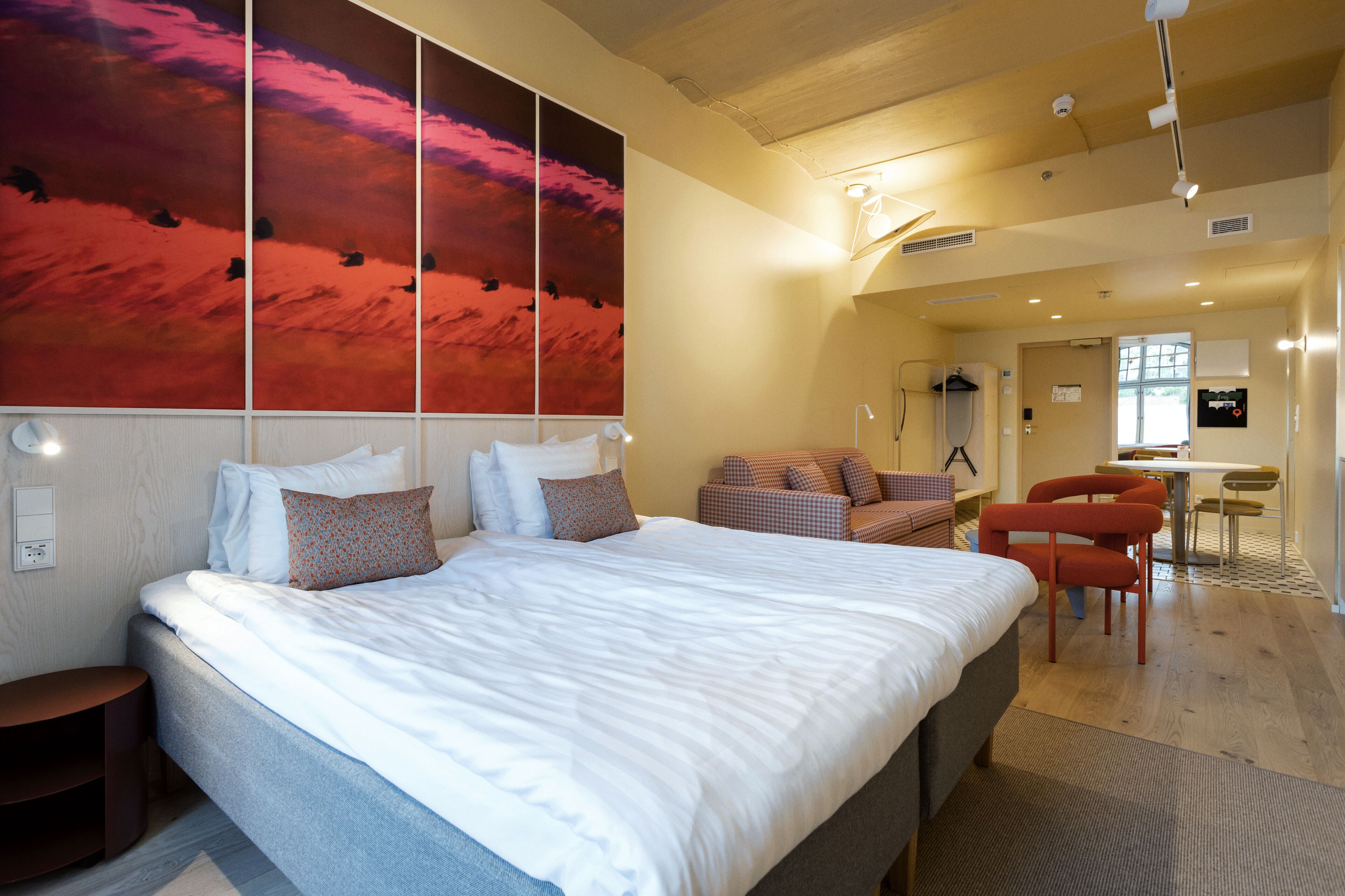 Quarto de hotel moderno com uma cama grande, área de estar com mesa e cadeiras, e uma pintura grande na parede.