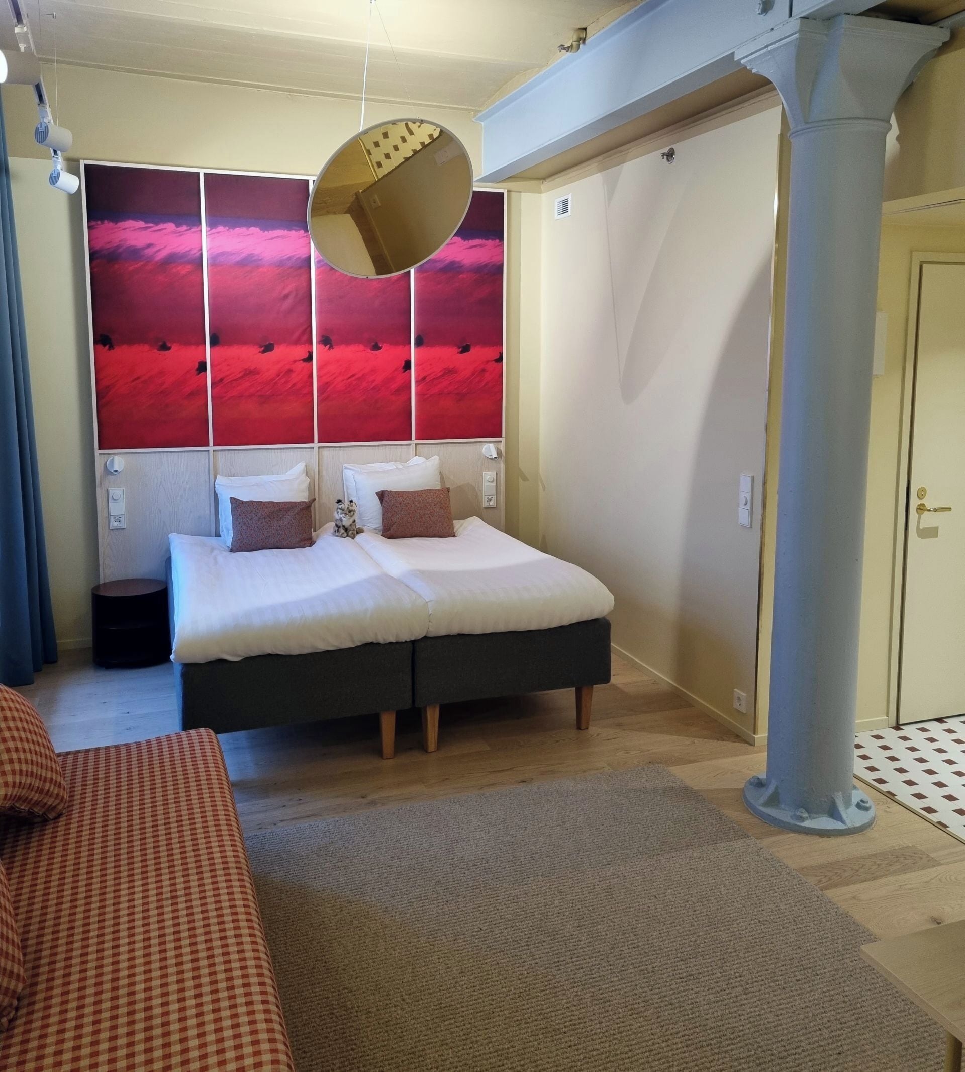 Quarto com cama, um espelho redondo pequeno, parede com arte vermelha, uma porta e uma janela.