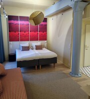 Quarto com cama, um espelho redondo pequeno, parede com arte vermelha, uma porta e uma janela.