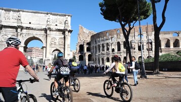 Sítio histórico com arcos e colunas antigas, incluindo o Arco de Tito e o Coliseu, com pessoas andando de bicicleta e caminhando.