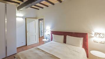 Quarto de hotel com cama, mesa de cabeceira, luminária na parede e porta para outro espaço.
