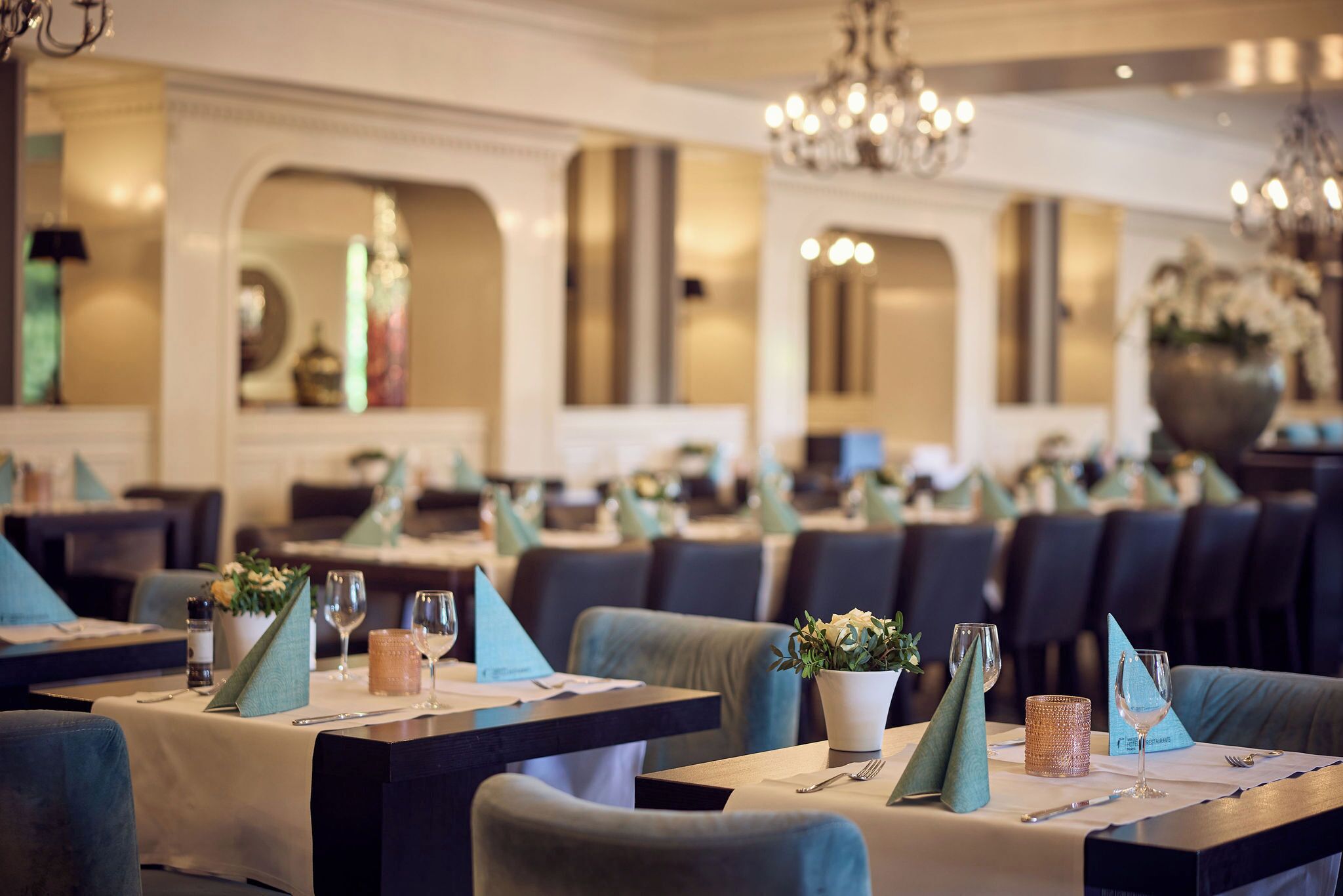 Interior de restaurante com mesas preparadas para refeições, cadeiras estofadas azuis, toalhas de mesa brancas e centros de mesa decorativos.