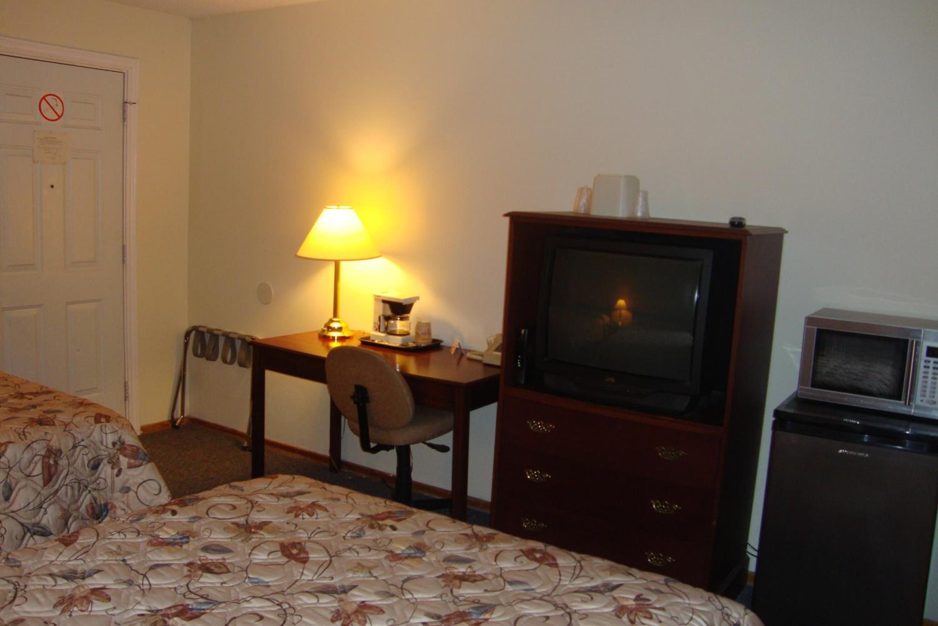 Quarto de hotel com cama, escrivaninha com luminária, televisão e refrigerador.