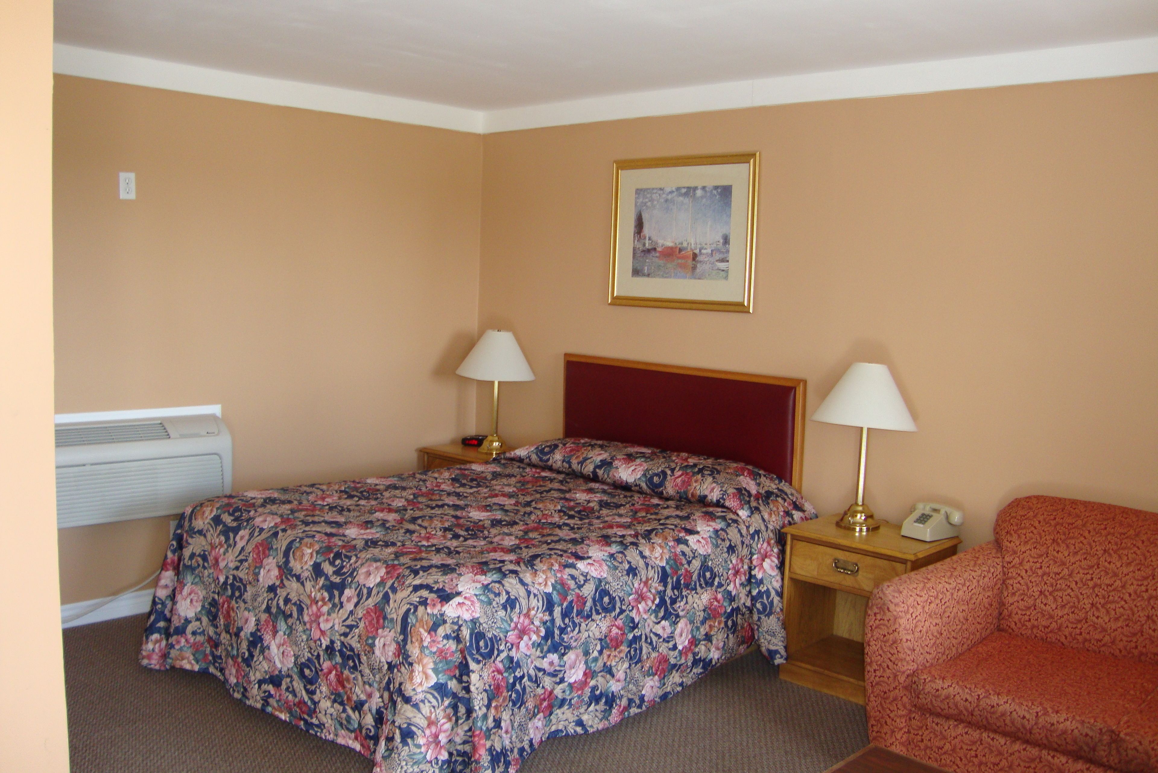 Quarto de hotel com uma cama, duas mesas de cabeceira, uma cadeira, um quadro na parede e um ar condicionado.