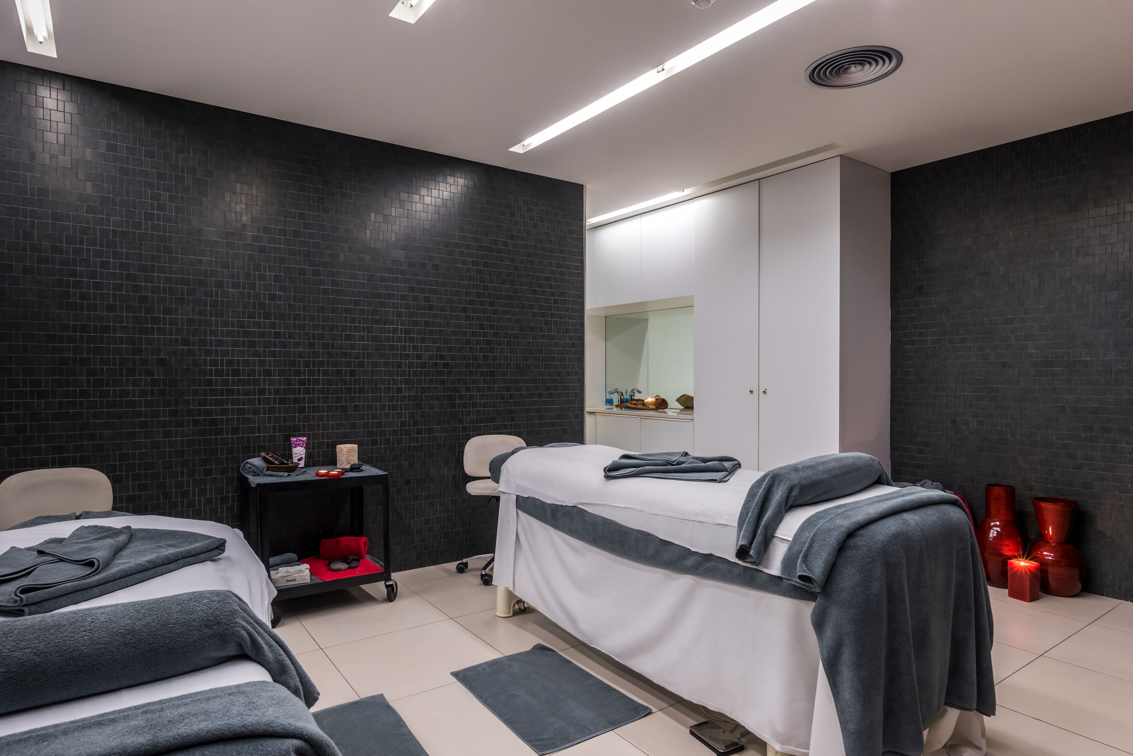 Uma sala de spa moderna com duas camas, uma mesa de massagem, uma pequena escrivaninha com artigos de higiene e um espelho.