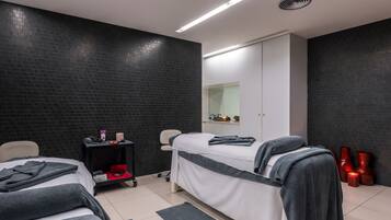 Uma sala de spa moderna com duas camas, uma mesa de massagem, uma pequena escrivaninha com artigos de higiene e um espelho.