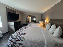 Quarto de hotel com uma cama grande, televisão fixada na parede, escrivaninha com abajur e uma porta em arco que dá para outro cômodo.