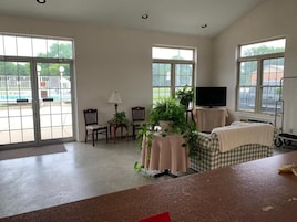 Quarto espaçoso com grandes janelas, televisão, mesa com planta e cadeira de rodas.