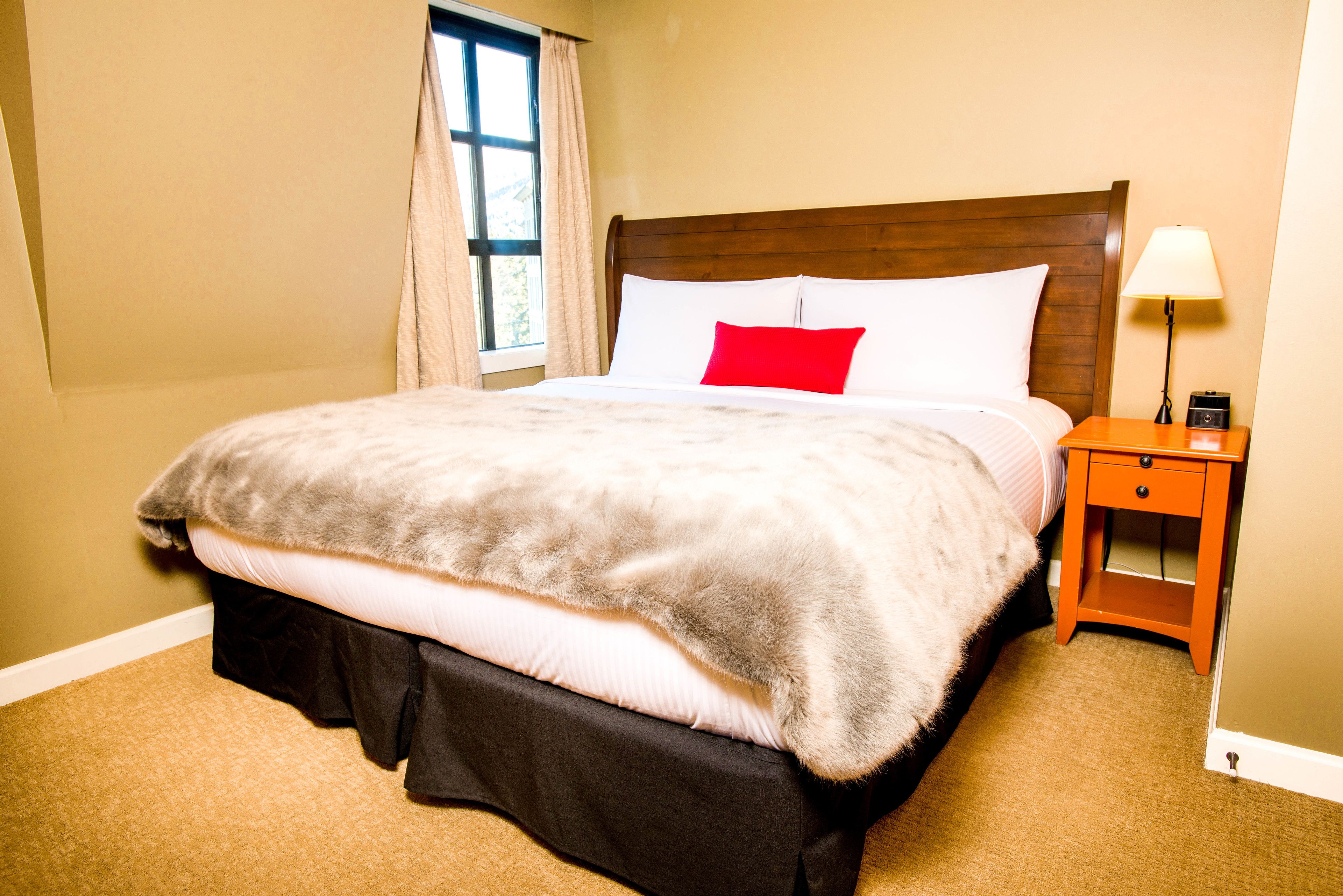 Cama bem arrumada com travesseiro branco e vermelho, cabeceira de madeira e mesa de cabeceira de madeira com uma luminária.