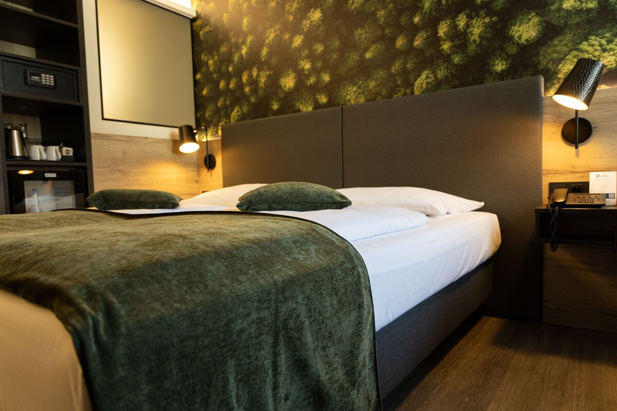 Quarto de hotel com cama, manta verde, mesa de cabeceira com abajur e parede com padrão de folhagem.