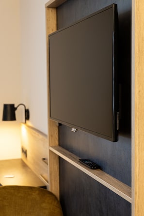Quarto moderno com televisão de tela plana fixada na parede, uma prateleira de madeira abaixo e um controle remoto sobre a prateleira.