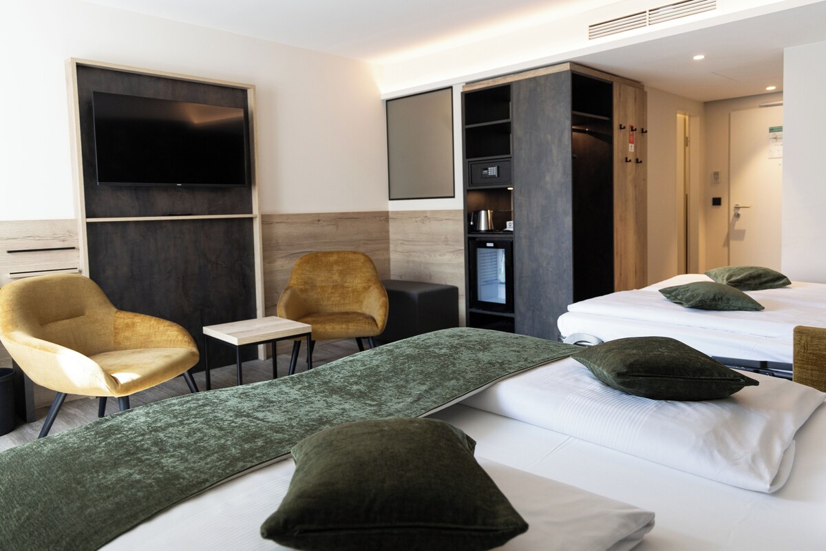 Quarto de hotel com duas camas, televisão, guarda-roupa e área de estar com cadeiras.