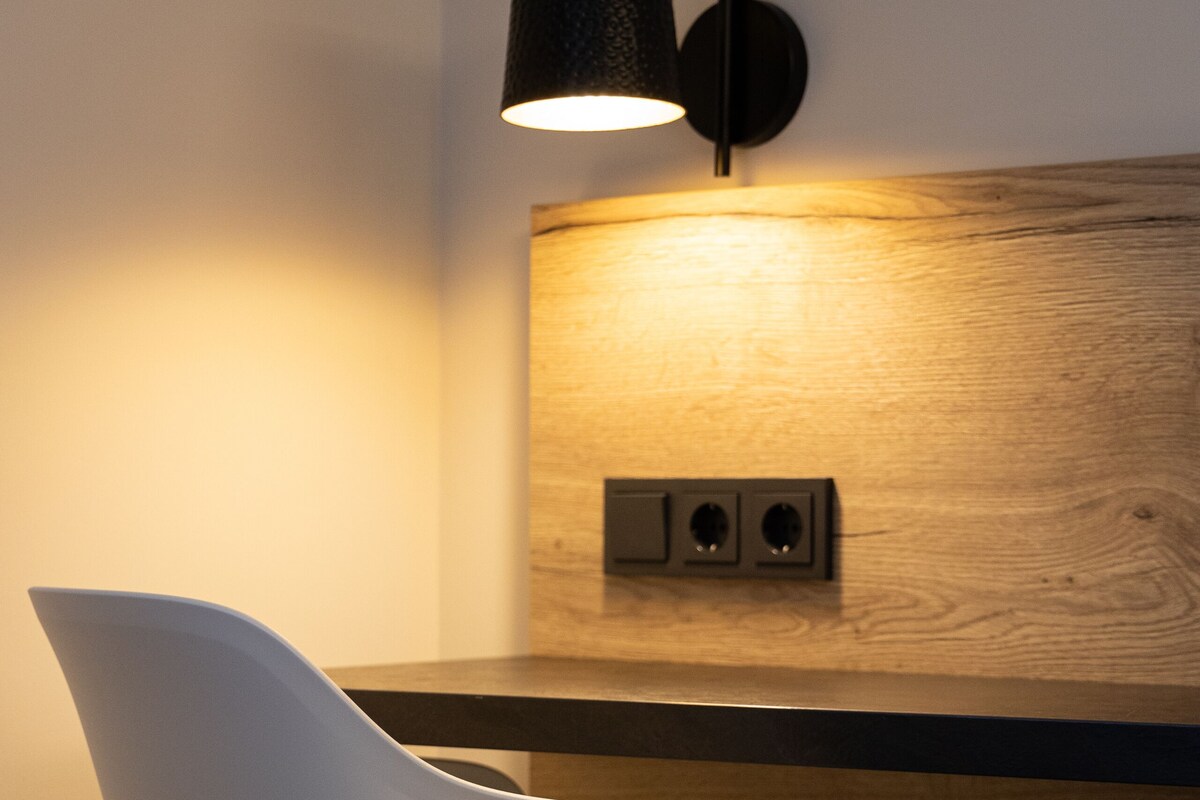 Uma cadeira moderna com assento branco e pés pretos, colocada ao lado de um balcão de madeira com uma luminária preta na parede.