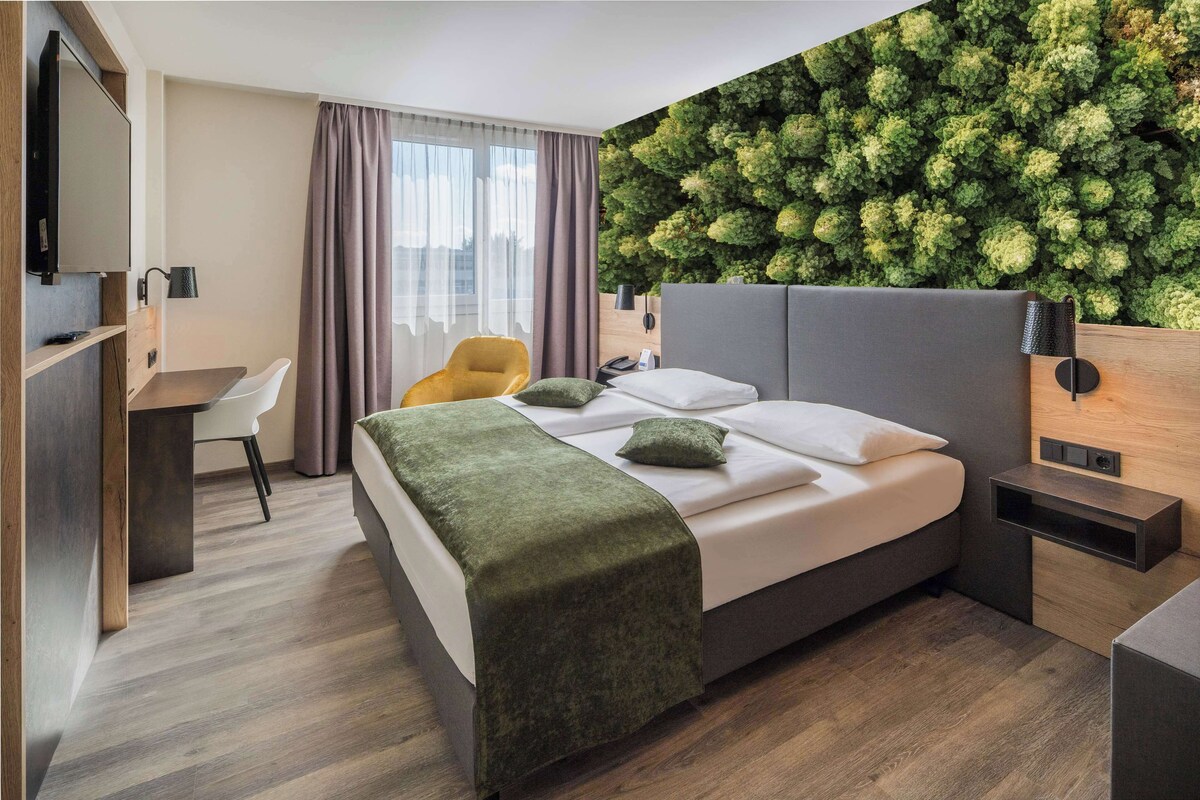 Quarto de hotel moderno com cama, escrivaninha, cadeira, TV e painel verde na parede.