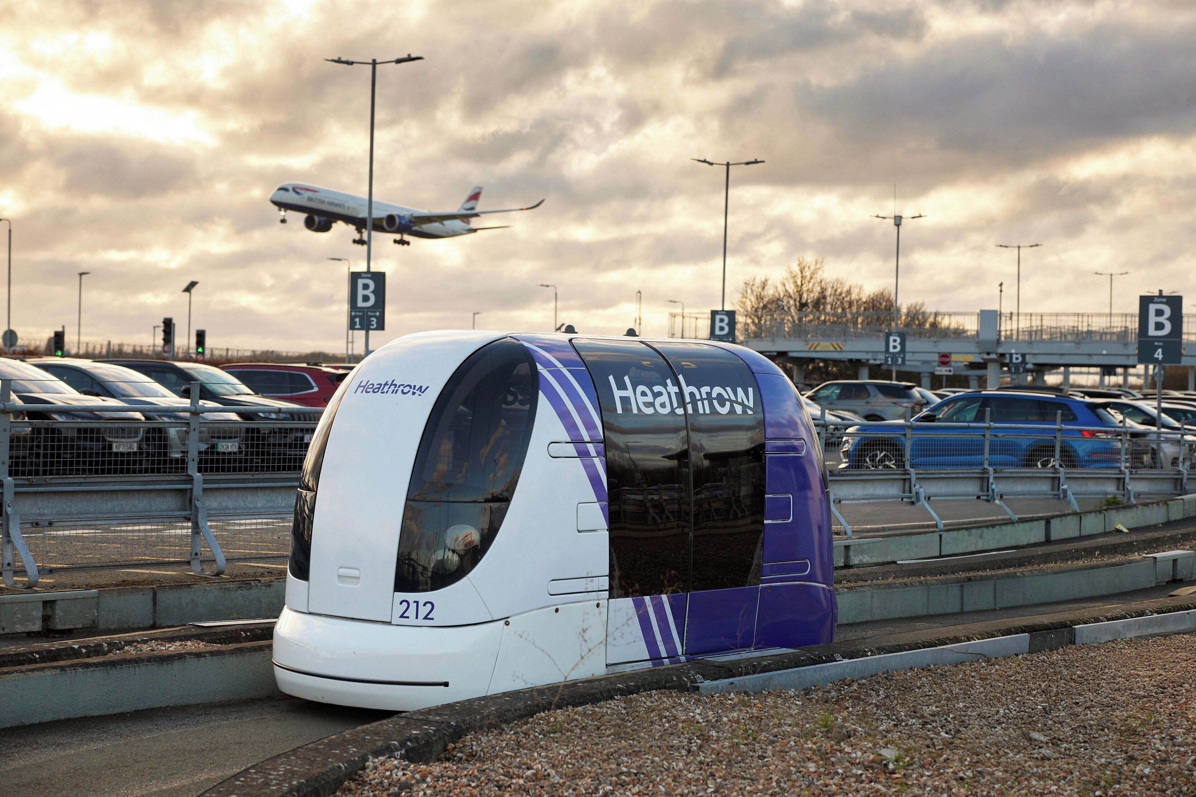 Um autocarro de transporte moderno, branco e roxo com a marca 'Heathrow', estacionado num terminal de aeroporto.