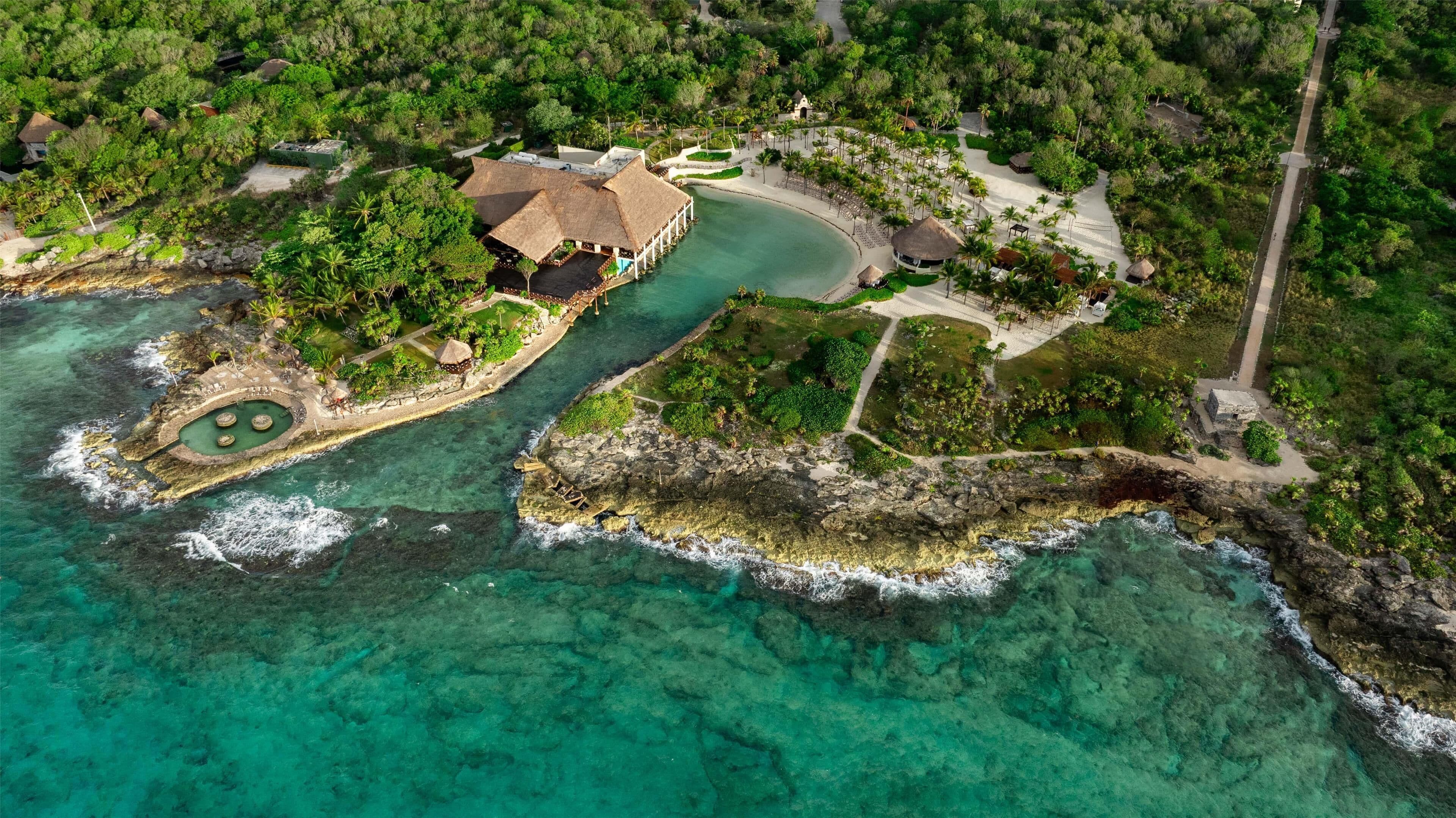 Vista aérea de um resort à beira-mar com praia, águas turquesa cristalinas e vegetação exuberante.
