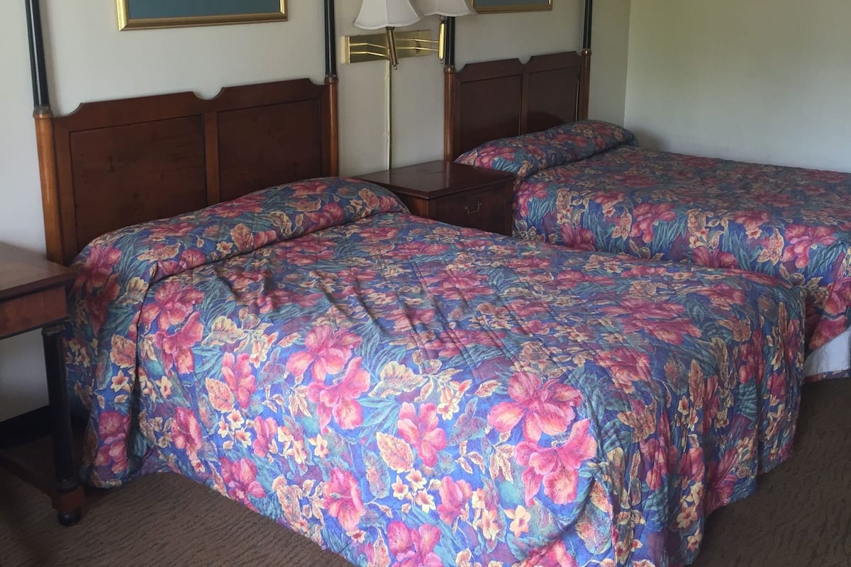 Quarto de hotel com duas camas, uma mesa de cabeceira, um abajur e duas fotos emolduradas na parede.