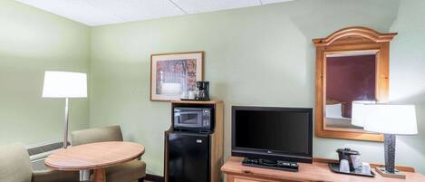 Quarto de hotel com uma mesa redonda de madeira, um sofá, um minibar, uma televisão de tela plana, uma cômoda de madeira e um espelho.