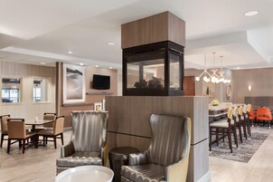 Um hall de hotel moderno com poltronas de couro, uma lareira central e uma área de refeições com cadeiras laranja.