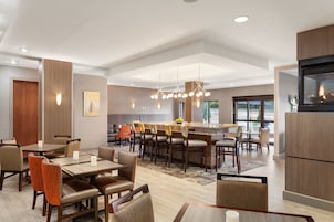 Uma zona de refeições moderna num hotel, com várias mesas e cadeiras, uma ilha central com balcão e uma área de cozinha visível à direita.