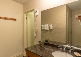 Casa de banho com bancada de granito, lavatório branco, secador de cabelo e espelho com parede de tijolos ao fundo.