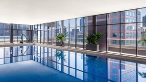 Uma grande piscina interior com paredes de vidro transparente, plantas em vasos e vista para a cidade.