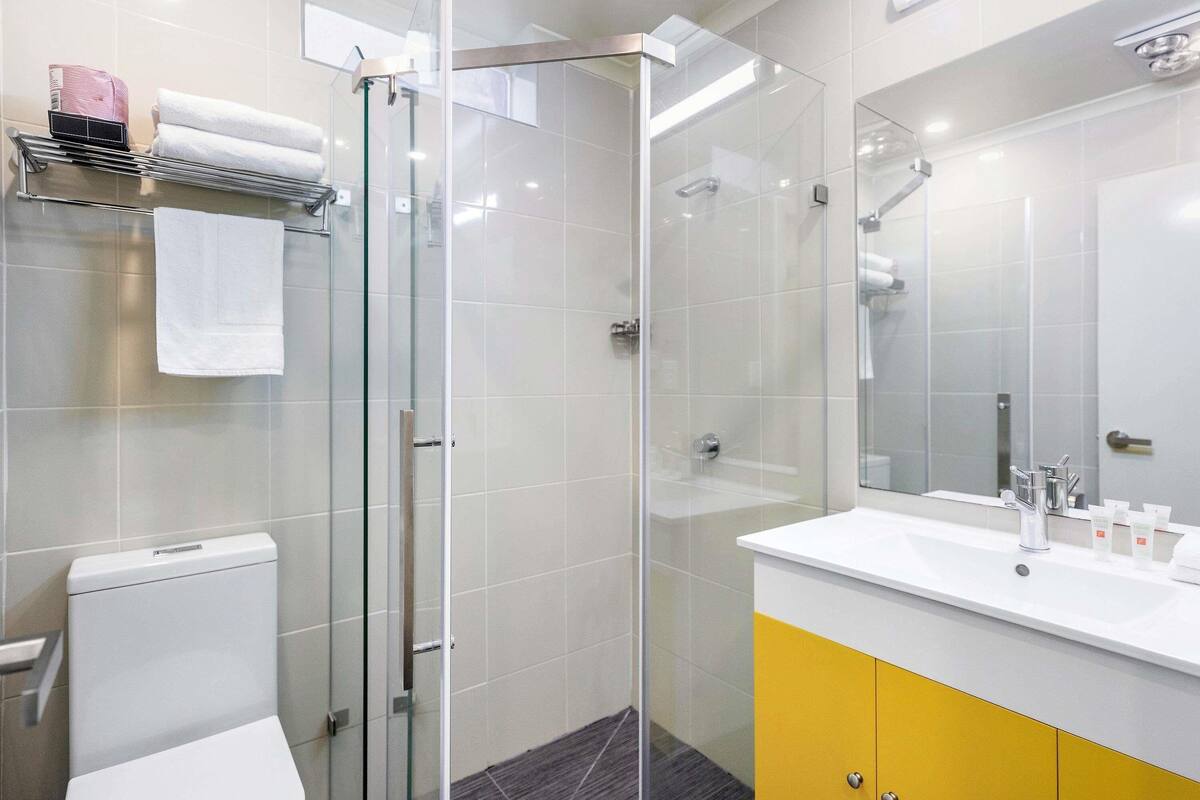 Uma casa de banho moderna com cabine de duche em vidro, sanita branca, toucador amarelo e branco e um espelho grande.