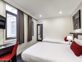 Quarto de hotel com duas camas, uma escrivaninha, uma cadeira, uma televisão e uma janela com cortinas.