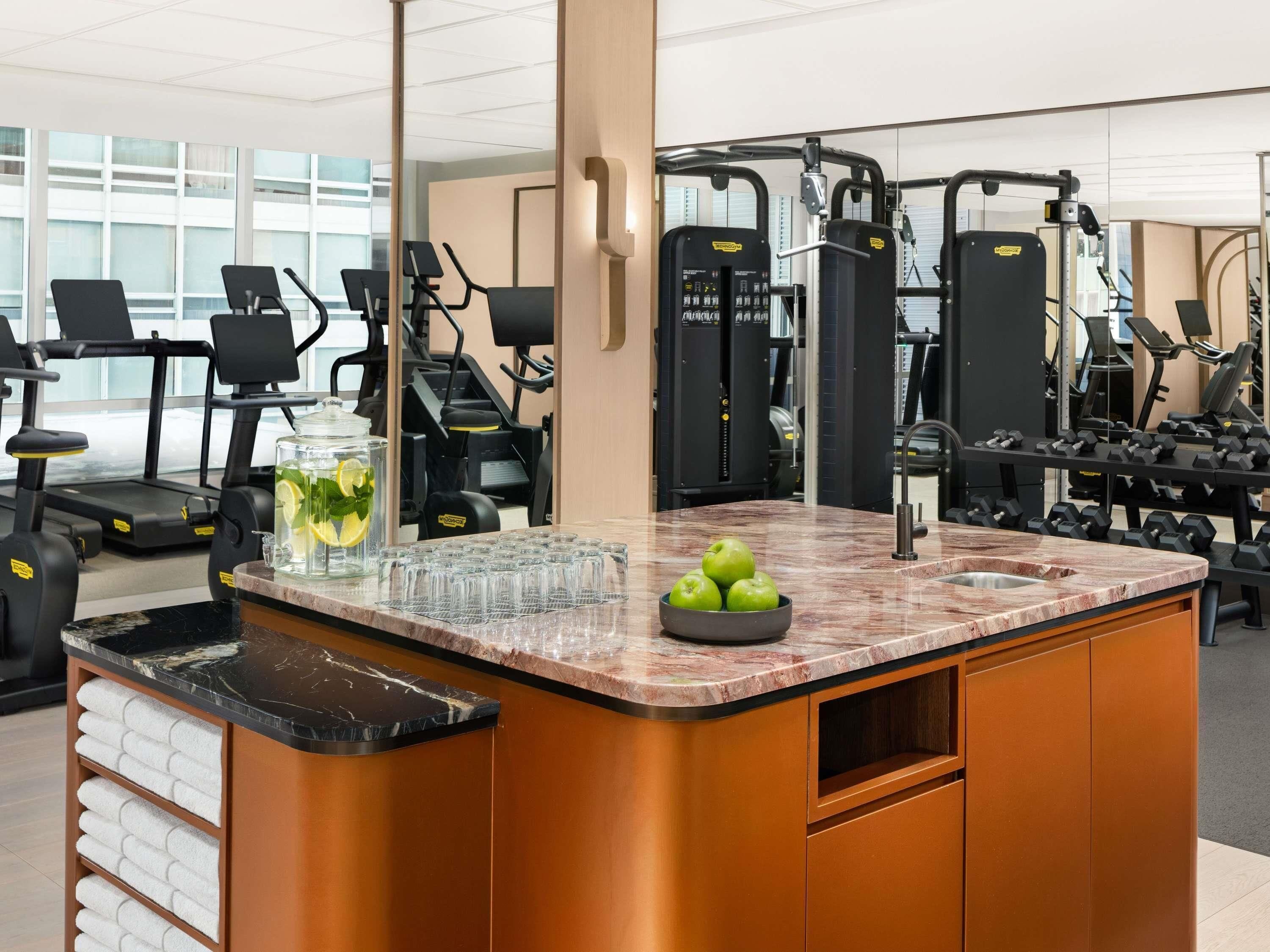 Uma academia com equipamentos de ginástica, um ponto de água e um balcão com uma tigela de frutas e copos.
