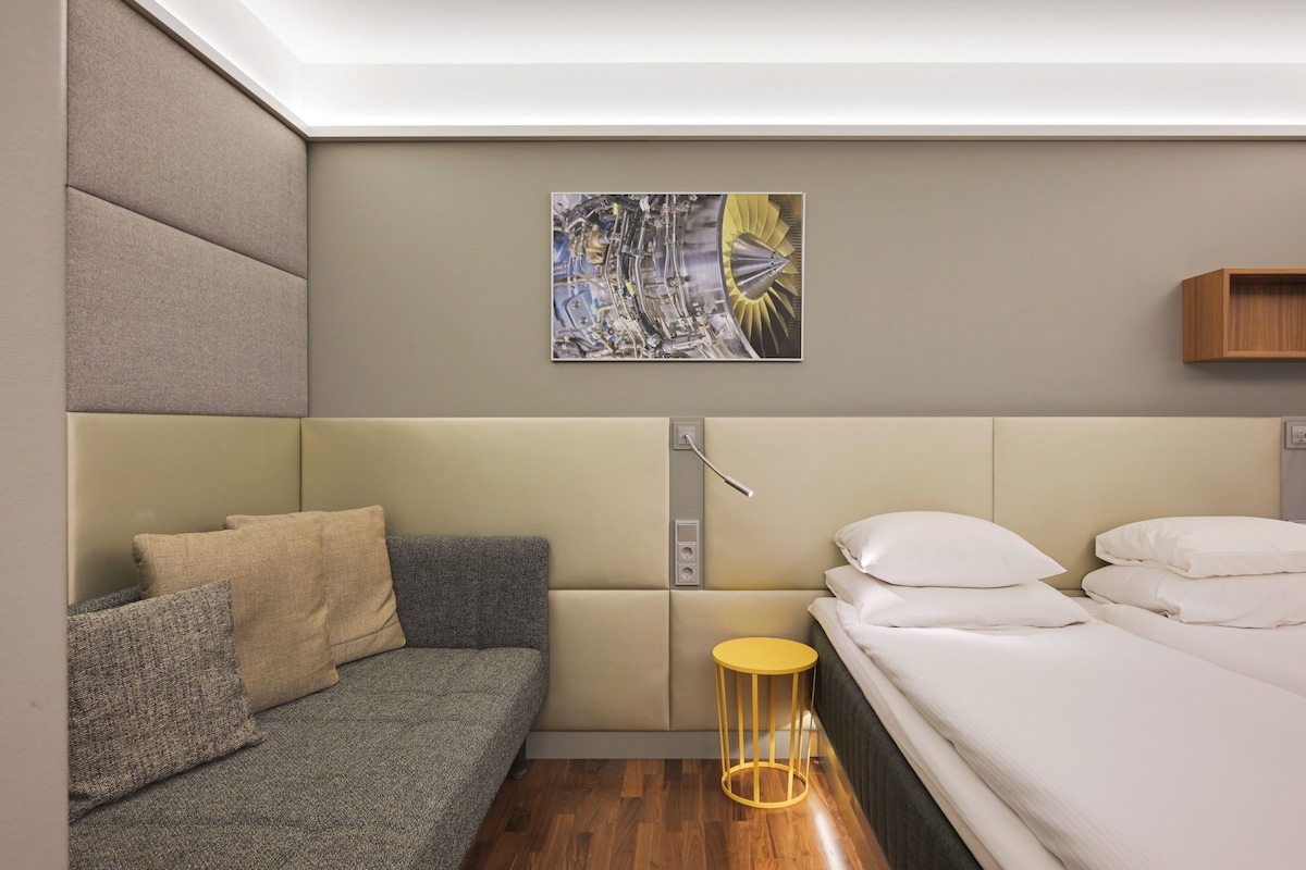 Quarto de hotel moderno com sofá, cama, mesa redonda pequena e uma obra de arte na parede.