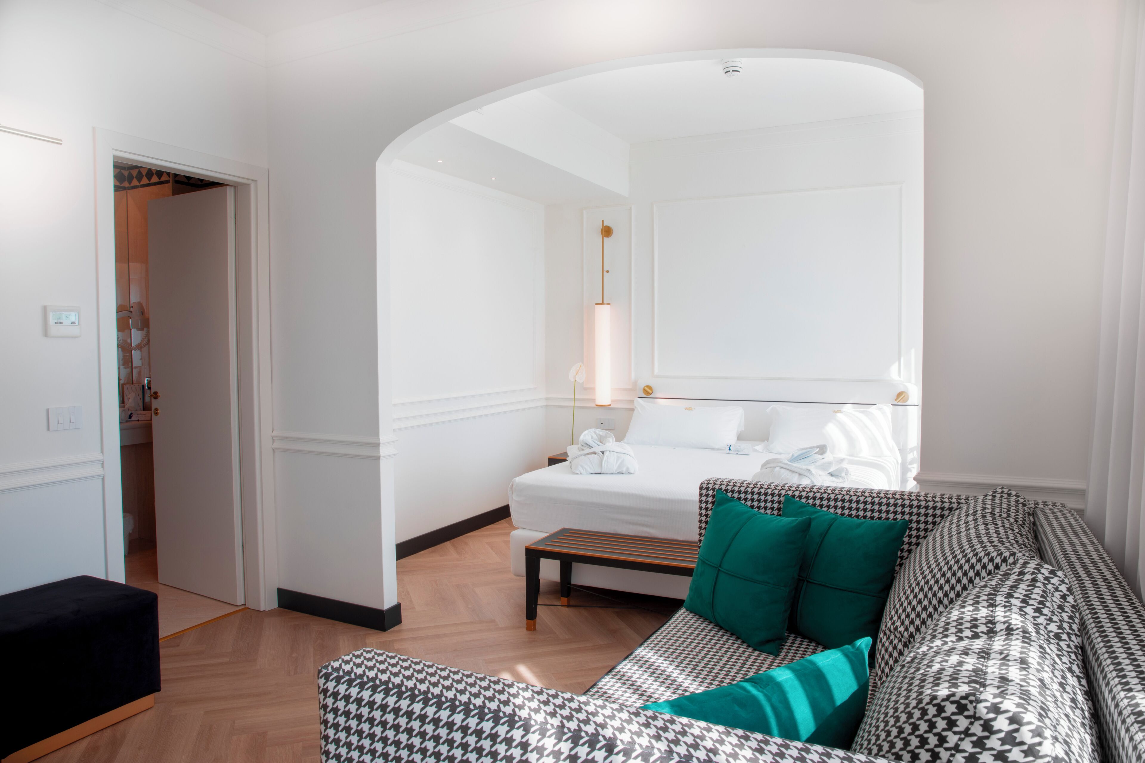 Quarto de hotel com cama, sofá xadrez, banco de madeira e parede branca.