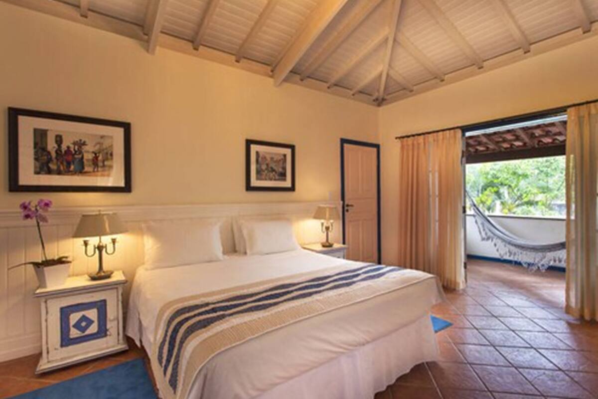 Um quarto com uma cama grande, uma mesinha de cabeceira com abajur, duas imagens emolduradas na parede e vista para o exterior através de uma porta.