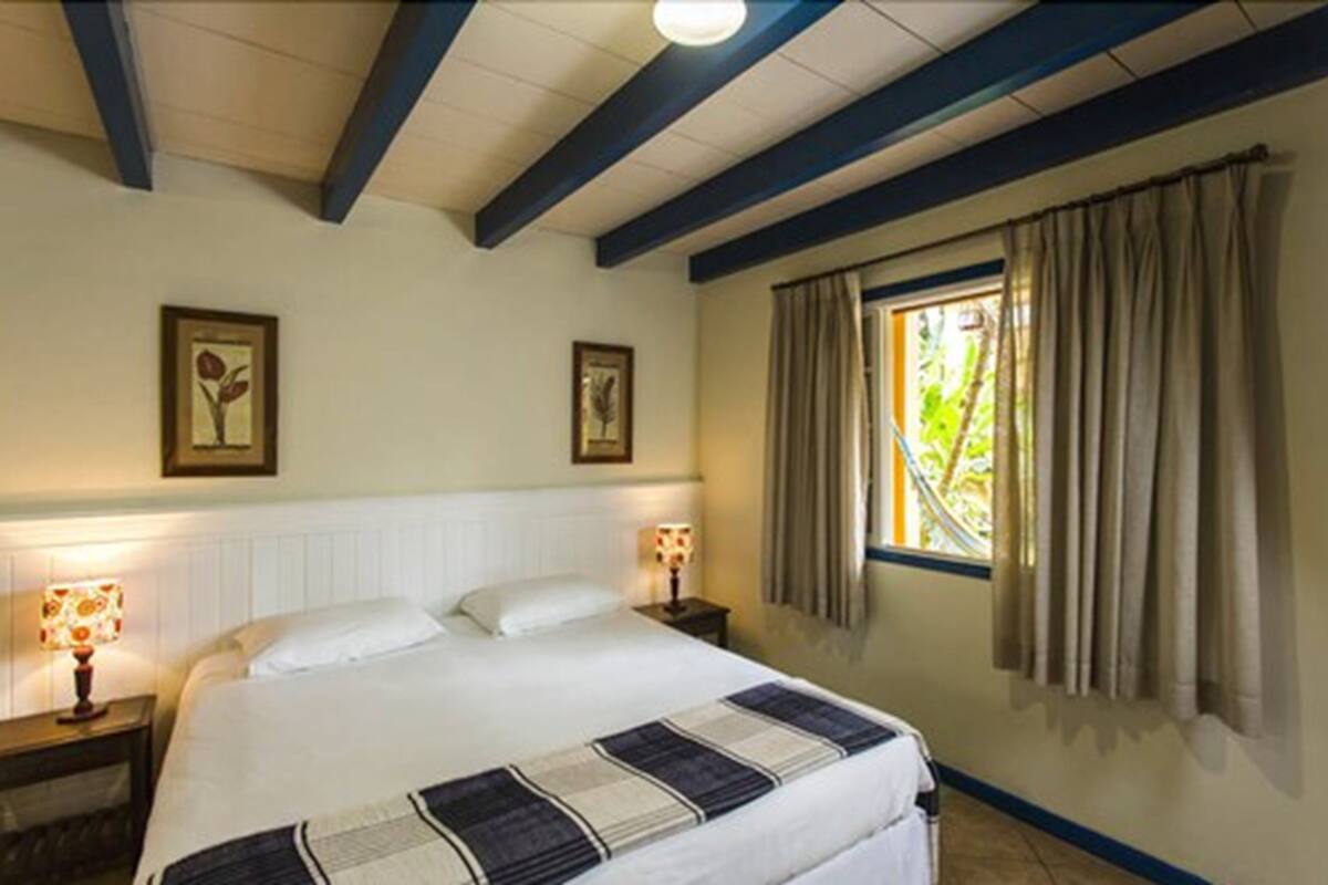 Quarto de hotel com uma cama grande, duas mesas de cabeceira com abajures, uma janela com cortinas e quadros nas paredes.
