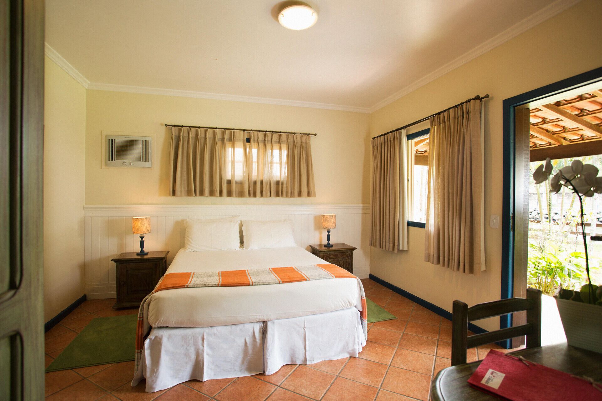 Quarto de hotel com cama, duas mesas de cabeceira, janela com cortinas, radiador e vista para o exterior.