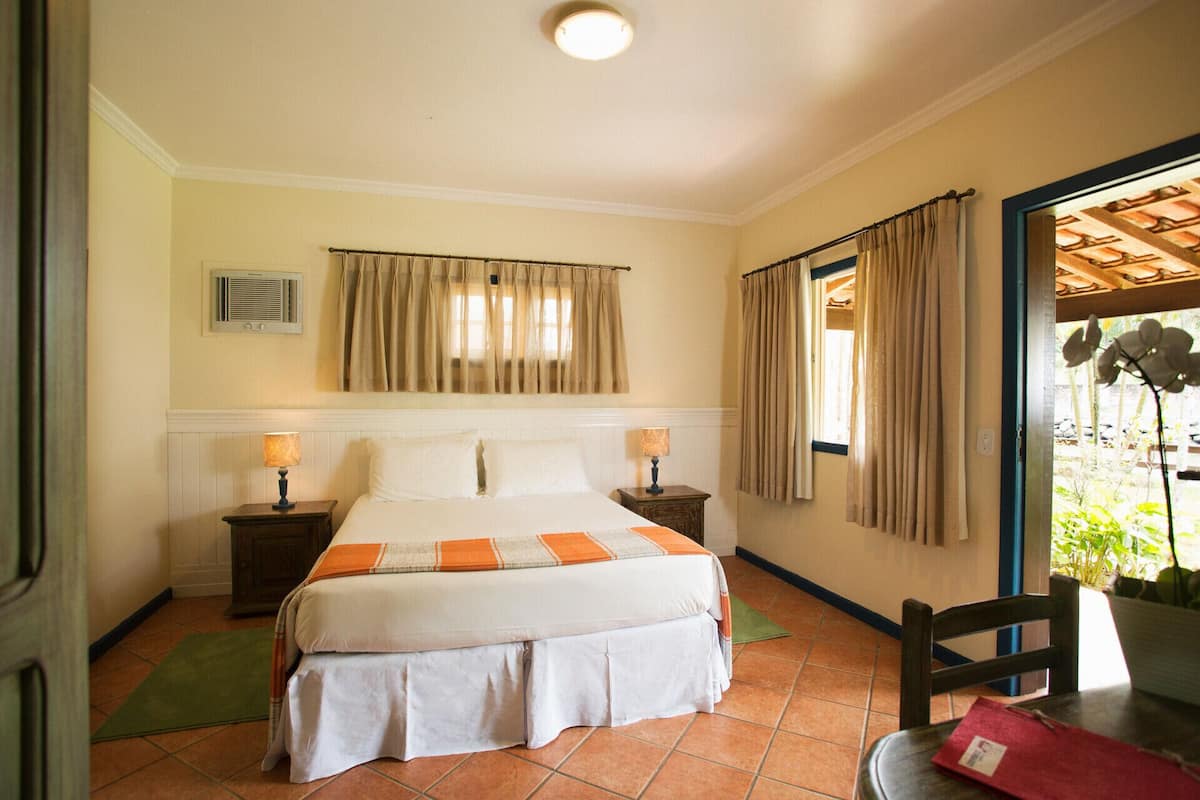 Quarto de hotel com cama, duas mesas de cabeceira, janela com cortinas, radiador e vista para o exterior.