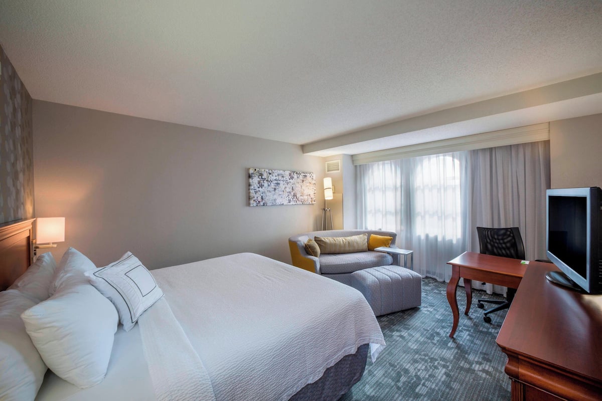 Quarto de hotel com uma cama grande, um sofá, uma escrivaninha com televisão e uma janela com cortinas.