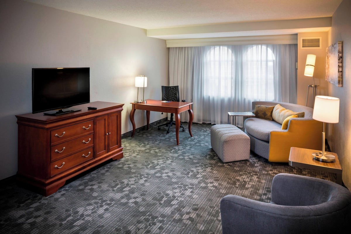 Quarto de hotel com televisão de tela plana, penteadeira de madeira, escrivaninha, cadeira de escritório, sofá, banquinho e abajur.