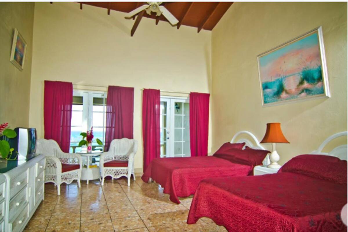 Quarto de hotel com duas camas, área de refeições com mesa e cadeiras, e ventilador de teto.