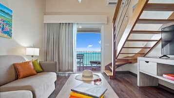 Quarto de hotel moderno com sofá, mesa de centro com livros e chapéu, TV em um suporte e varanda com vista para o mar.