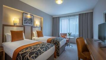 Quarto de hotel com duas camas, uma mesa, uma cadeira, uma televisão e uma janela com cortinas.