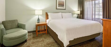 Quarto de hotel com cama, poltrona, mesa de cabeceira, abajur e janela com cortinas.