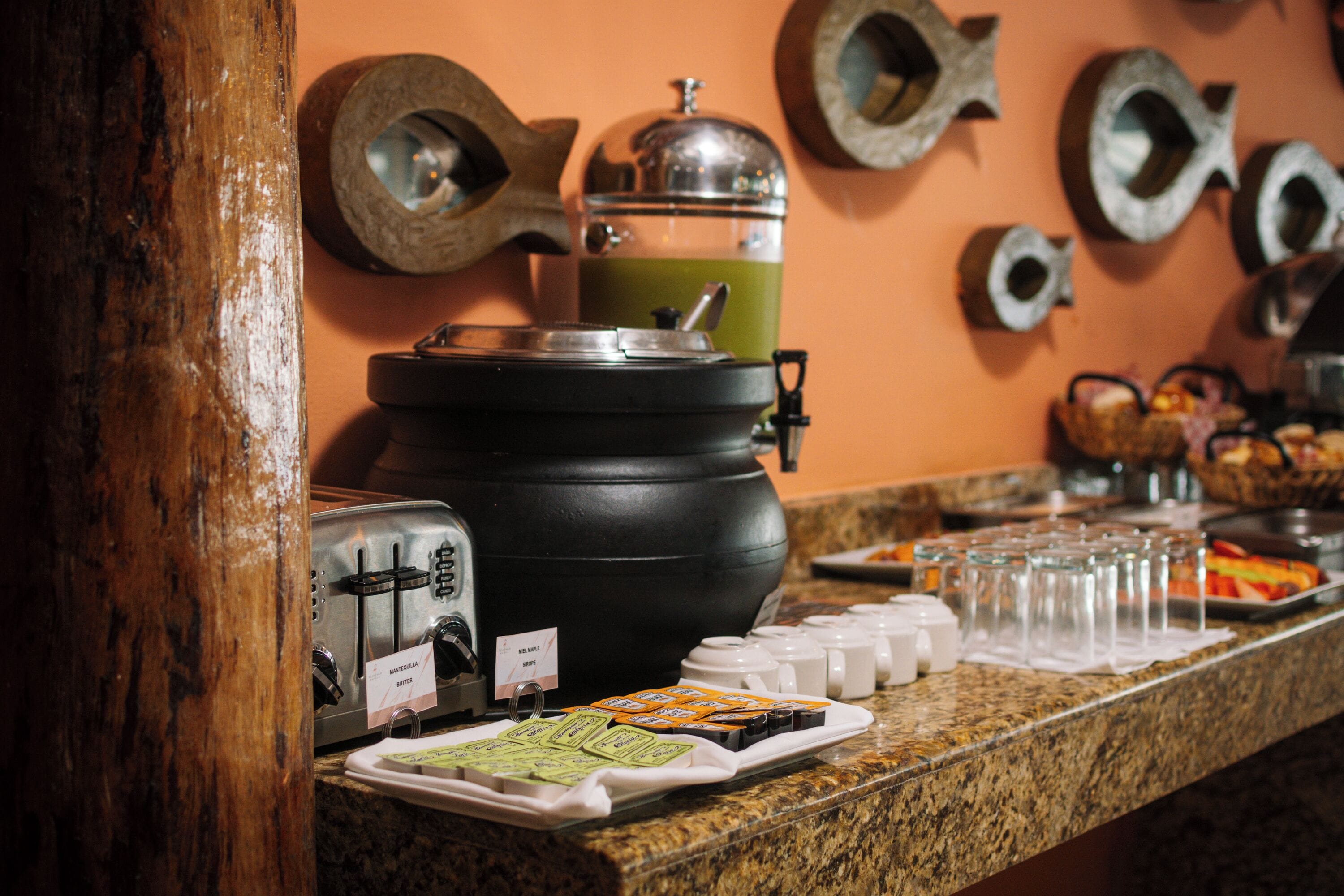 Disposição de buffet com uma cafeteira, uma panela grande e diversos alimentos sobre um balcão de granito.