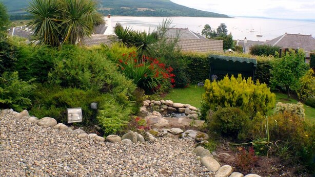 Um jardim com diversas plantas, um caminho de pedra e vista para um corpo de água e montanhas ao longe.