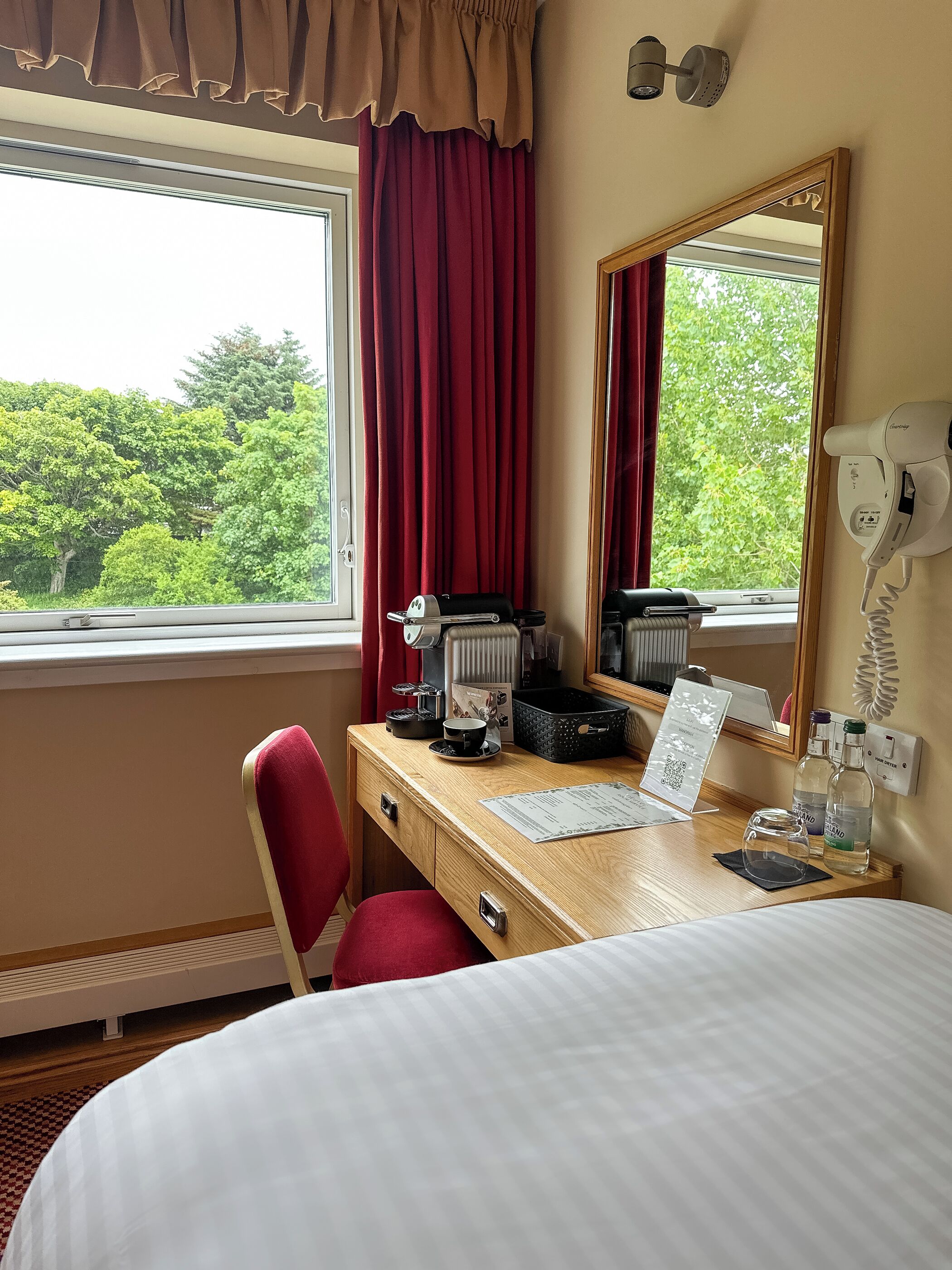 Quarto de hotel com uma escrivaninha de madeira, uma cadeira vermelha, uma cama com roupa de cama branca e uma janela com cortinas vermelhas, oferecendo vista para árvores.