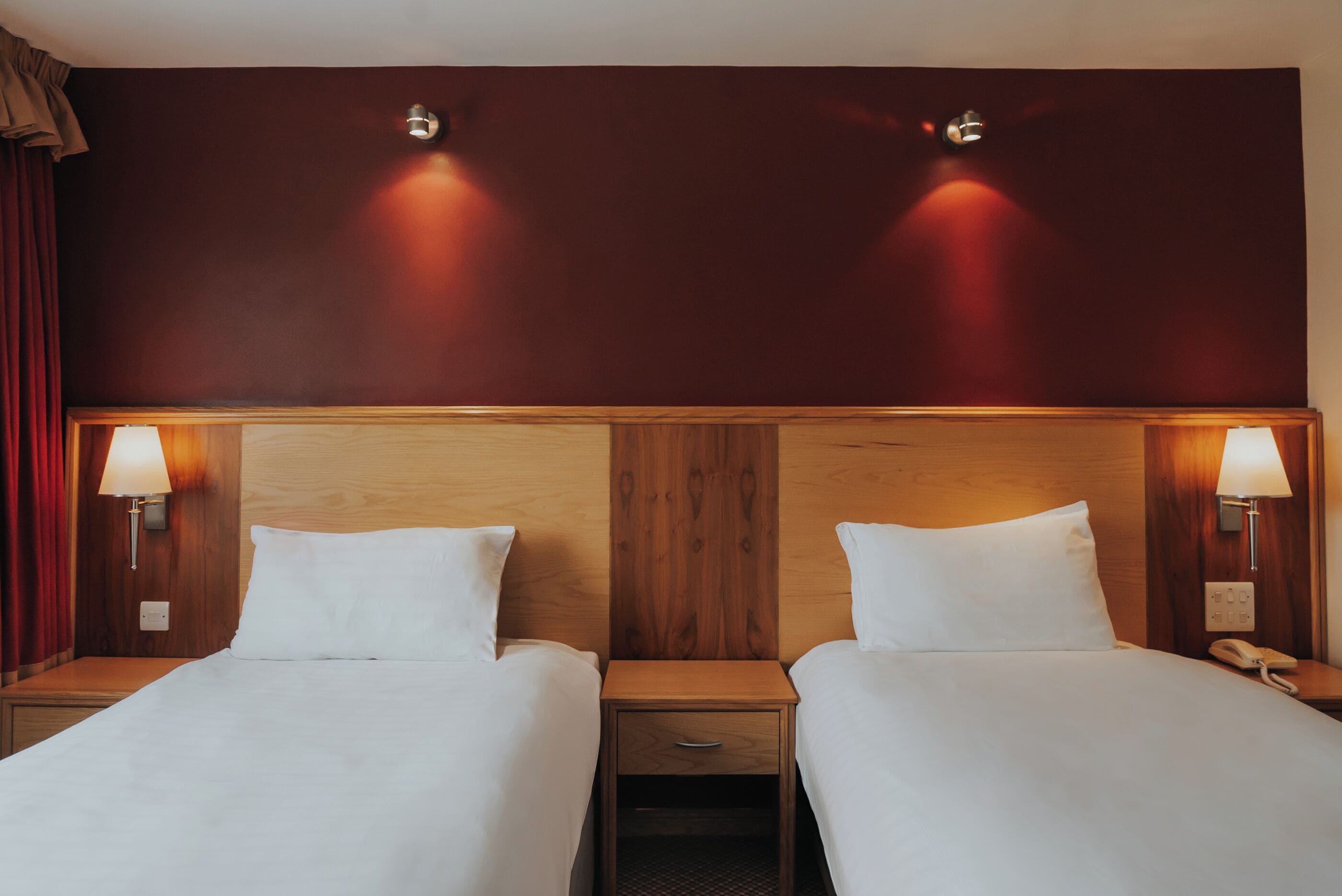 Quarto de hotel com duas camas, cabeceira de madeira, abajures e uma pequena gaveta.