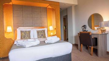 Quarto de hotel com uma cama grande, duas luminárias de cabeceira, uma escrivaninha com cadeira, um espelho redondo e um telefone fixado na parede.