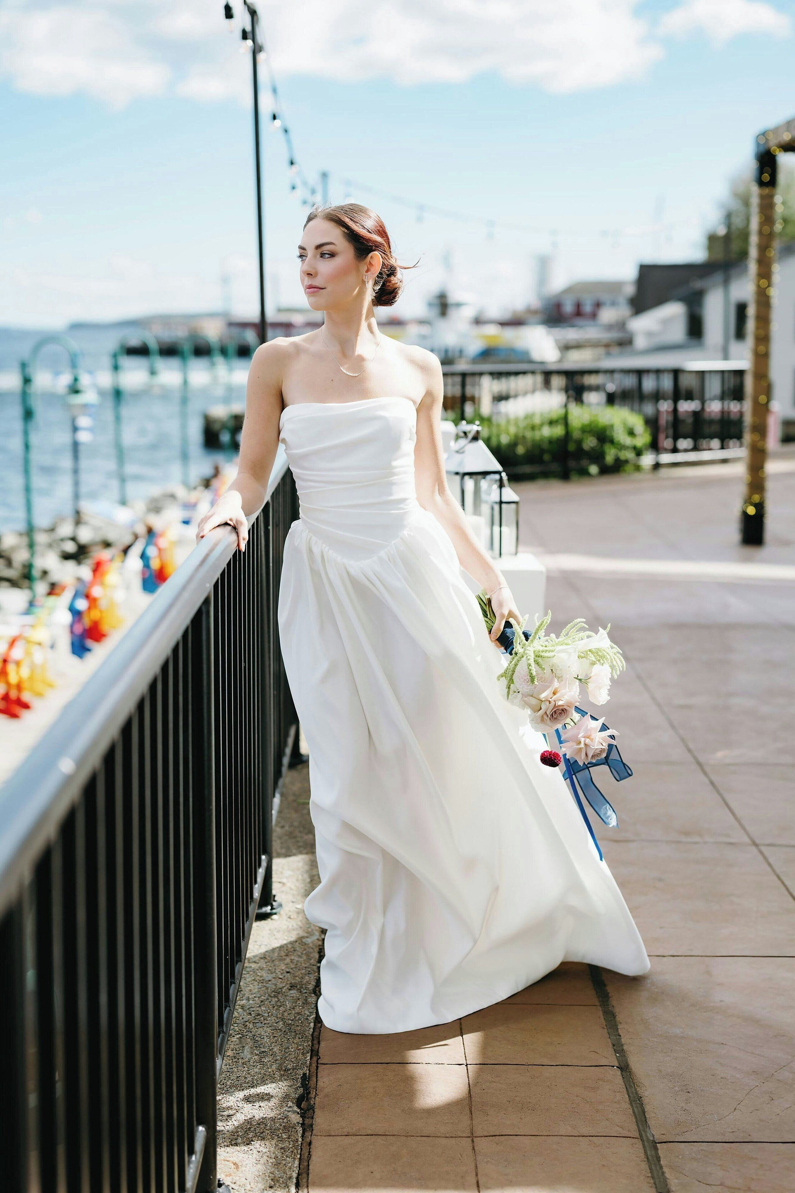 Uma pessoa de vestido branco de casamento parada junto a um corrimão, com vista para a água e edifícios ao fundo.