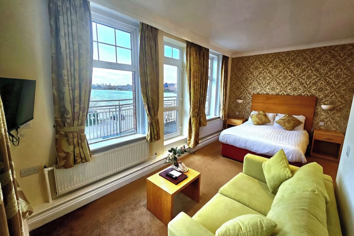 Quarto de hotel com cama, sofá verde, mesa de cabeceira de madeira, televisão e janela com cortinas.