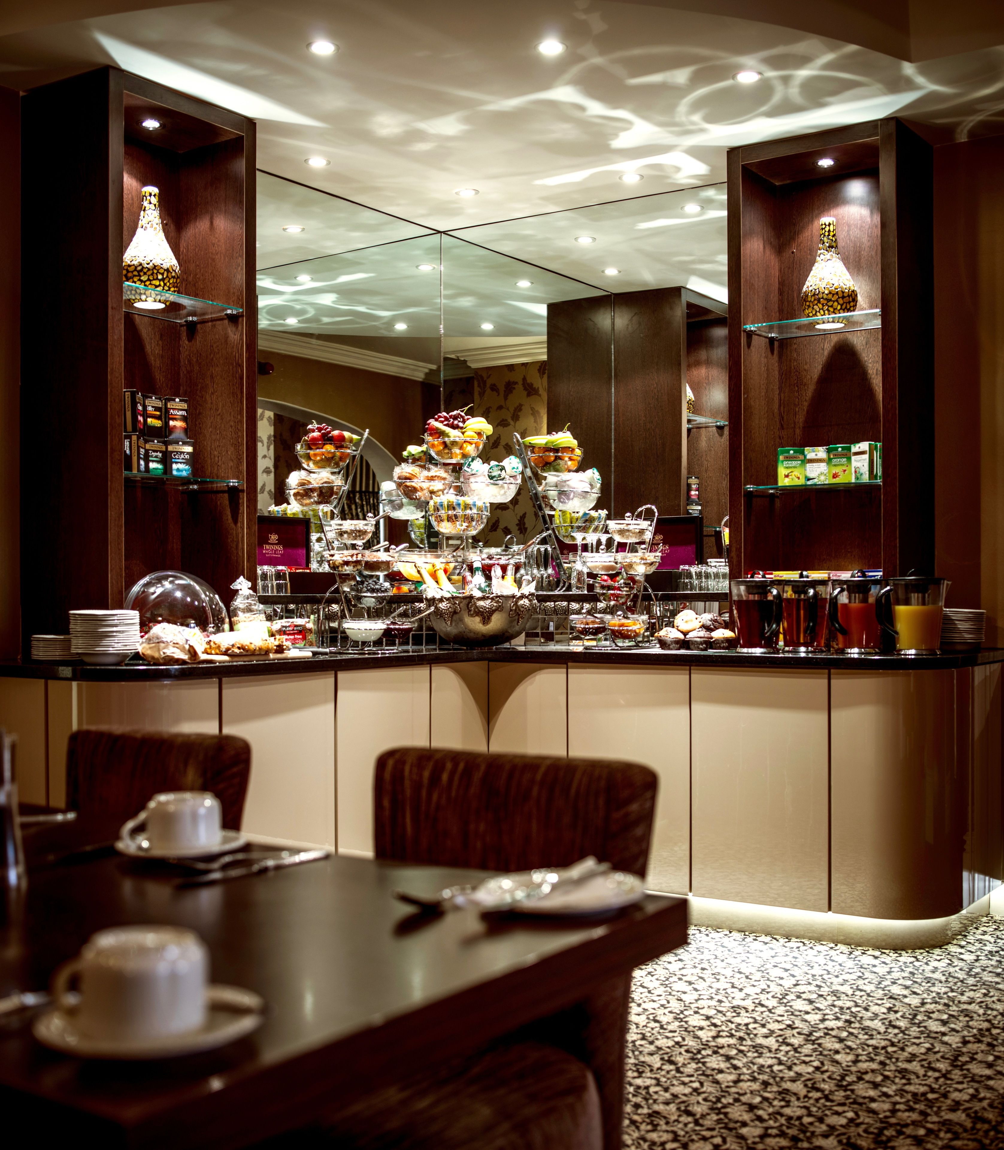 Um buffet de café da manhã com diversos alimentos e bebidas, uma área de estar com mesas e cadeiras, e um teto decorativo com iluminação embutida.